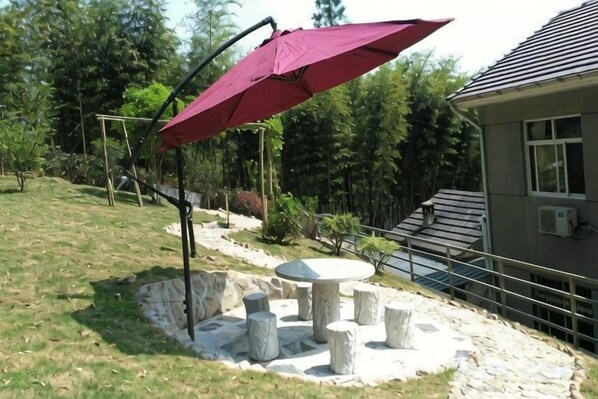 Uma área de estar externa com mesa e cadeiras, uma pérgula com um guarda-sol vermelho e uma parede de pedra.
