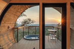 Um deck de madeira com uma banheira de hidromassagem, uma mesa e cadeiras, com vista para uma paisagem deslumbrante ao pôr do sol.