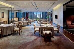 Ampla sala de jantar de hotel com mesas e cadeiras redondas, janelas grandes e vista para a paisagem urbana.