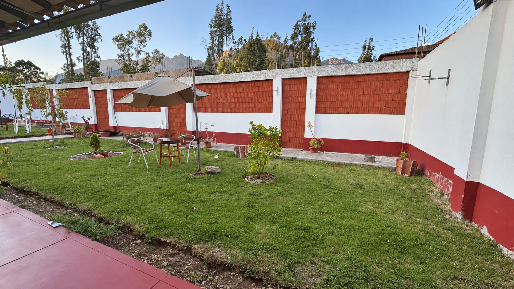 Um pátio com um edifício vermelho e branco, uma área de estar coberta e plantas em vasos.
