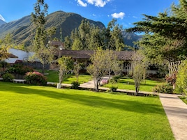 Um jardim bem cuidado com um caminho que leva a um edifício com telhado em cerâmica, cercado por vegetação exuberante e montanhas ao fundo.