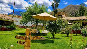 Um sinal de madeira indica direções para um restaurante, recepção e bar, situado em um jardim exuberante, com montanhas ao fundo.