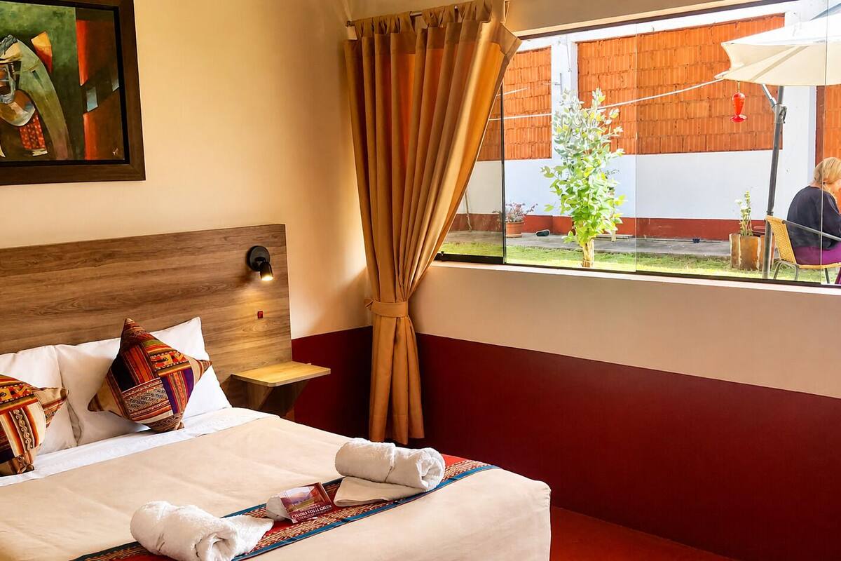 Quarto de hotel com cama, almofadas, mesa de cabeceira, janela com cortinas e um quadro na parede.