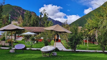 Um jardim com mesas ao ar livre, entrada de um edifício e montanhas ao fundo.
