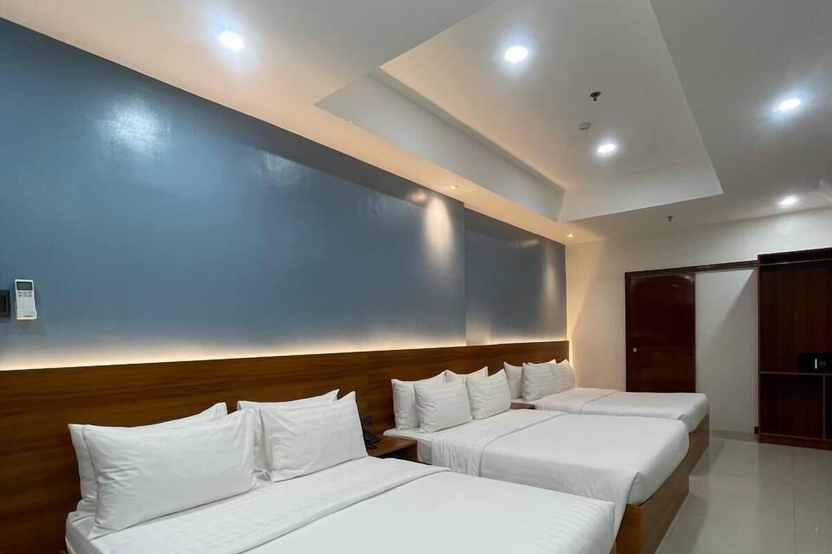 Quarto de hotel com três camas, cabeceira de madeira, parede grande com detalhe azul e iluminação embutida no teto.