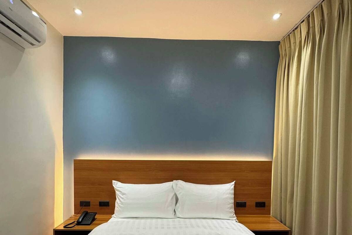 Quarto de hotel com cama, cabeceira de madeira, mesa de cabeceira e cortina.