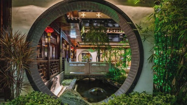 Elemento arquitetônico circular com um corpo d'água e vegetação, cercado por edifícios de estilo tradicional.