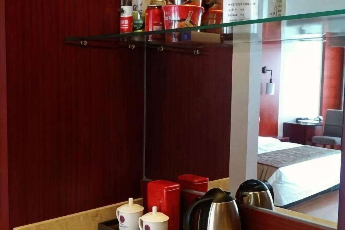 Quarto de hotel com divisória de vidro, cama, escrivaninha e parede vermelha.