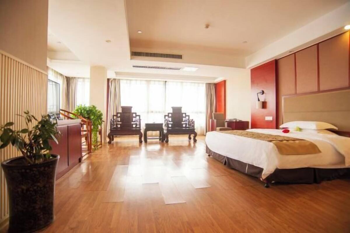 Quarto de hotel com cama, escrivaninha, cadeira, planta em vaso e janela com cortinas.
