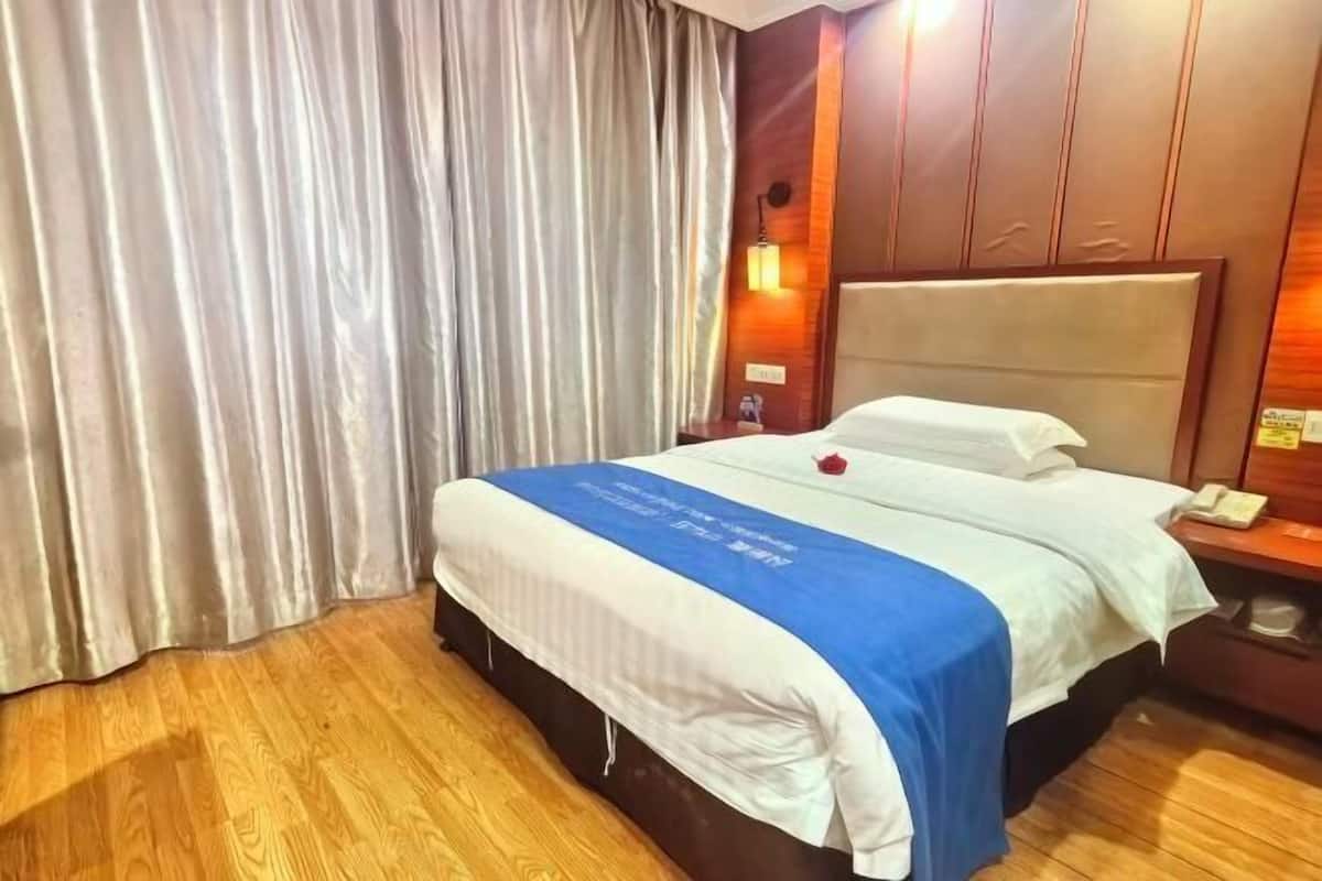 Quarto de hotel com cama, piso de madeira, cortinas, mesa de cabeceira e luminária na parede.