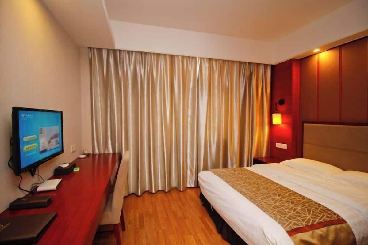 Quarto de hotel com uma escrivaninha de madeira, uma cama com colcha estampada, uma televisão na escrivaninha e uma cortina cobrindo a janela.