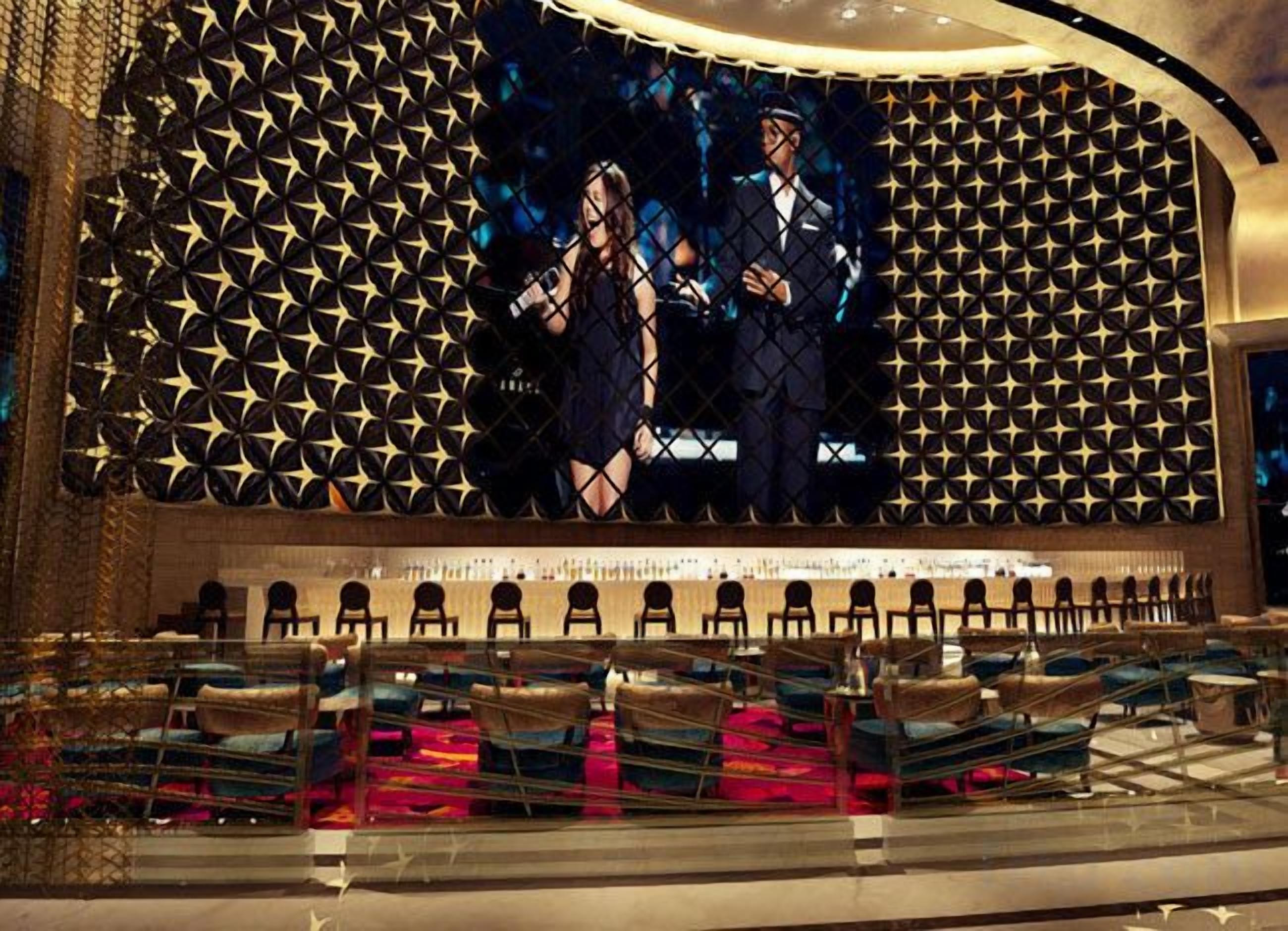 Um palco com um painel de fundo estampado com padrão geométrico, fileiras de cadeiras e uma área de bar com garrafas.
