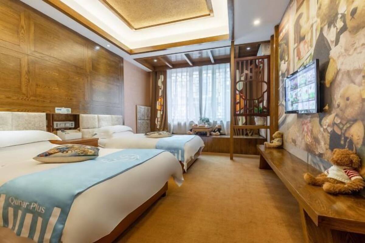 Quarto de hotel com duas camas, cabeceira de madeira, televisão, um urso de pelúcia e um mural na parede.