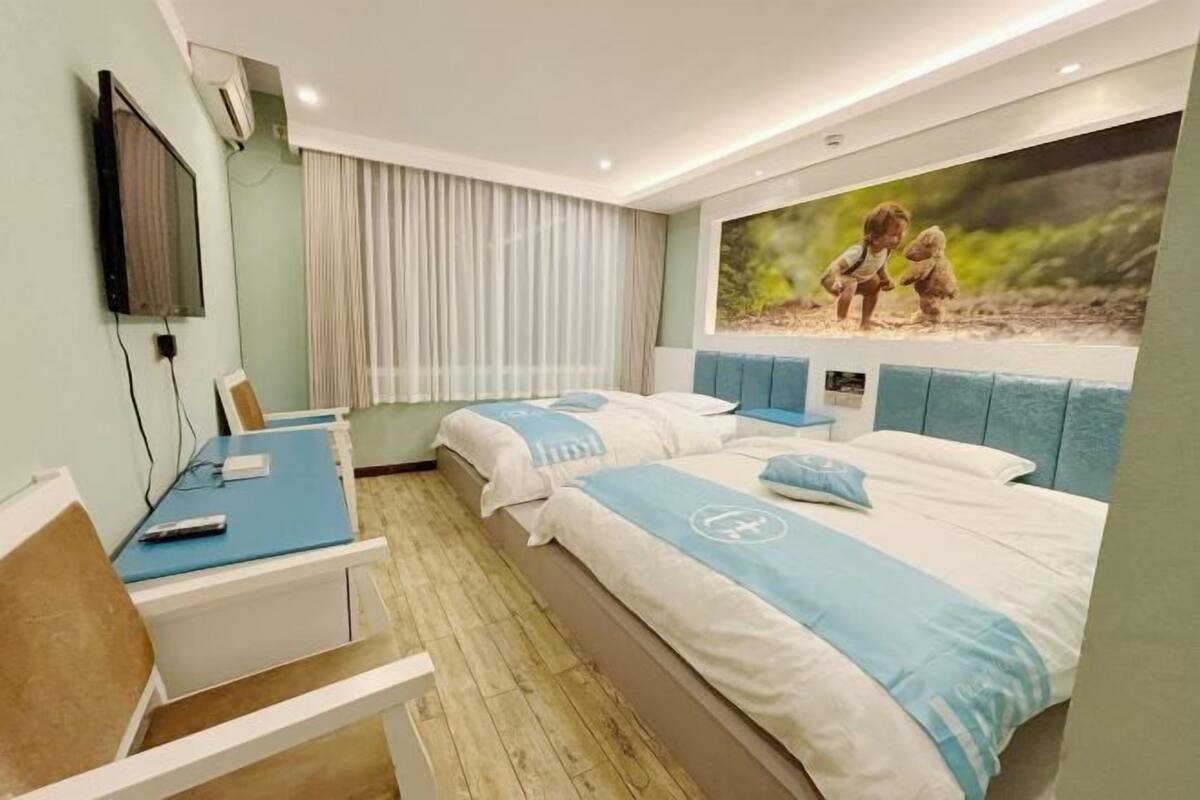 Quarto de hotel com duas camas, uma escrivaninha, televisão e uma janela grande com cortinas.