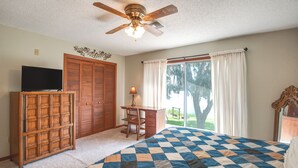 Quarto com uma cama grande, um guarda-roupa de madeira, uma mesa com cadeira, um ventilador de teto e uma janela com cortinas.