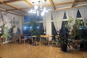 Uma área de refeições com mesas e cadeiras de madeira, plantas em vasos e grandes janelas com cortinas.