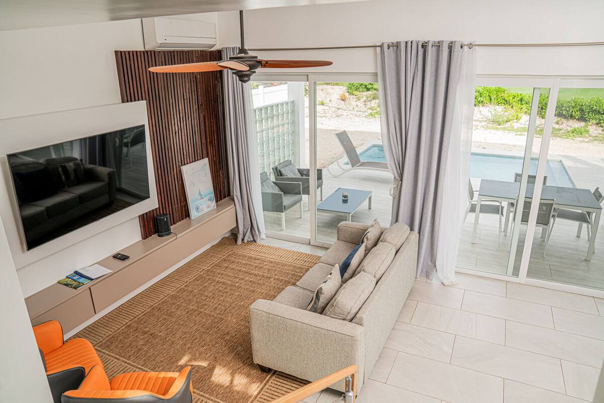 Uma sala de estar moderna com uma TV de tela plana, um ventilador de teto e uma grande porta de vidro deslizante que dá para uma área externa com piscina.