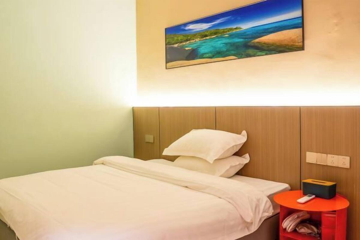 Quarto de hotel com cama, mesa de cabeceira, mesa lateral vermelha e pintura de uma praia.