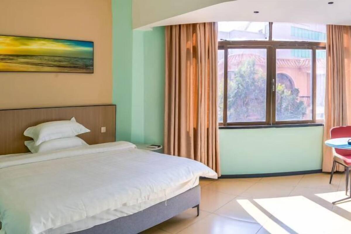 Quarto de hotel com uma cama, janela com cortinas, uma cadeira e um quadro na parede.