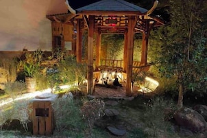 Um gazebo de madeira com cobertura, rodeado por um jardim com diversas plantas e um caminho que leva até ele.
