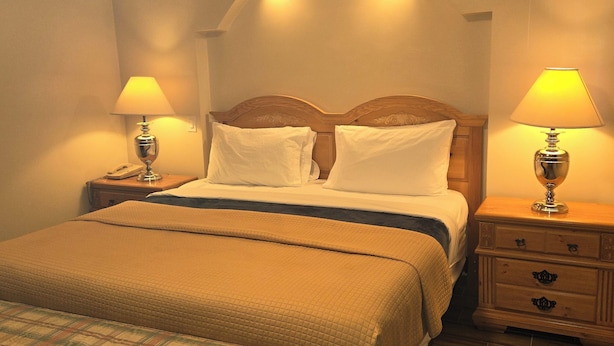 Quarto de hotel com uma cama grande, duas mesas de cabeceira com abajures, um banco e piso de madeira.