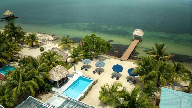 Vista aérea de um resort com piscina, guarda-sóis de praia, cadeiras de descanso, palmeiras e um píer que se estende para o oceano.