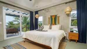 Quarto com uma cama grande, uma mesa de cabeceira de madeira, um tapete tecido e vista para palmeiras pelas portas da varanda.