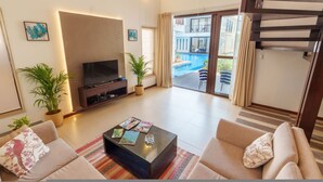 Uma sala de estar moderna com uma TV de tela plana, dois sofás, uma mesa de centro com revistas e uma grande janela com vista para uma piscina.