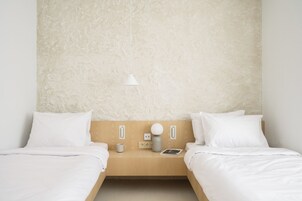 Quarto de hotel minimalista com duas camas de solteiro, cabeceira de madeira, mesa de cabeceira com uma lâmpada, uma chávena e um livro.