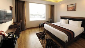 Quarto de hotel com uma cama grande, uma mesa com cadeira, televisão fixada na parede e uma janela ampla com vista para a cidade.