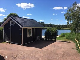 Uma cabana pequena e escura com telhado inclinado, uma janela e unidade de ar condicionado, localizada em área de cascalho com um lago e vegetação ao fundo.