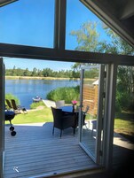 Varanda envidraçada com vista para um lago, móveis de exterior e uma pequena mesa com uma flor.