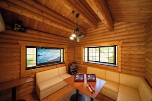 Uma cabana com paredes de madeira, duas janelas com vista, uma mesa com dispositivos eletrônicos e um banco.