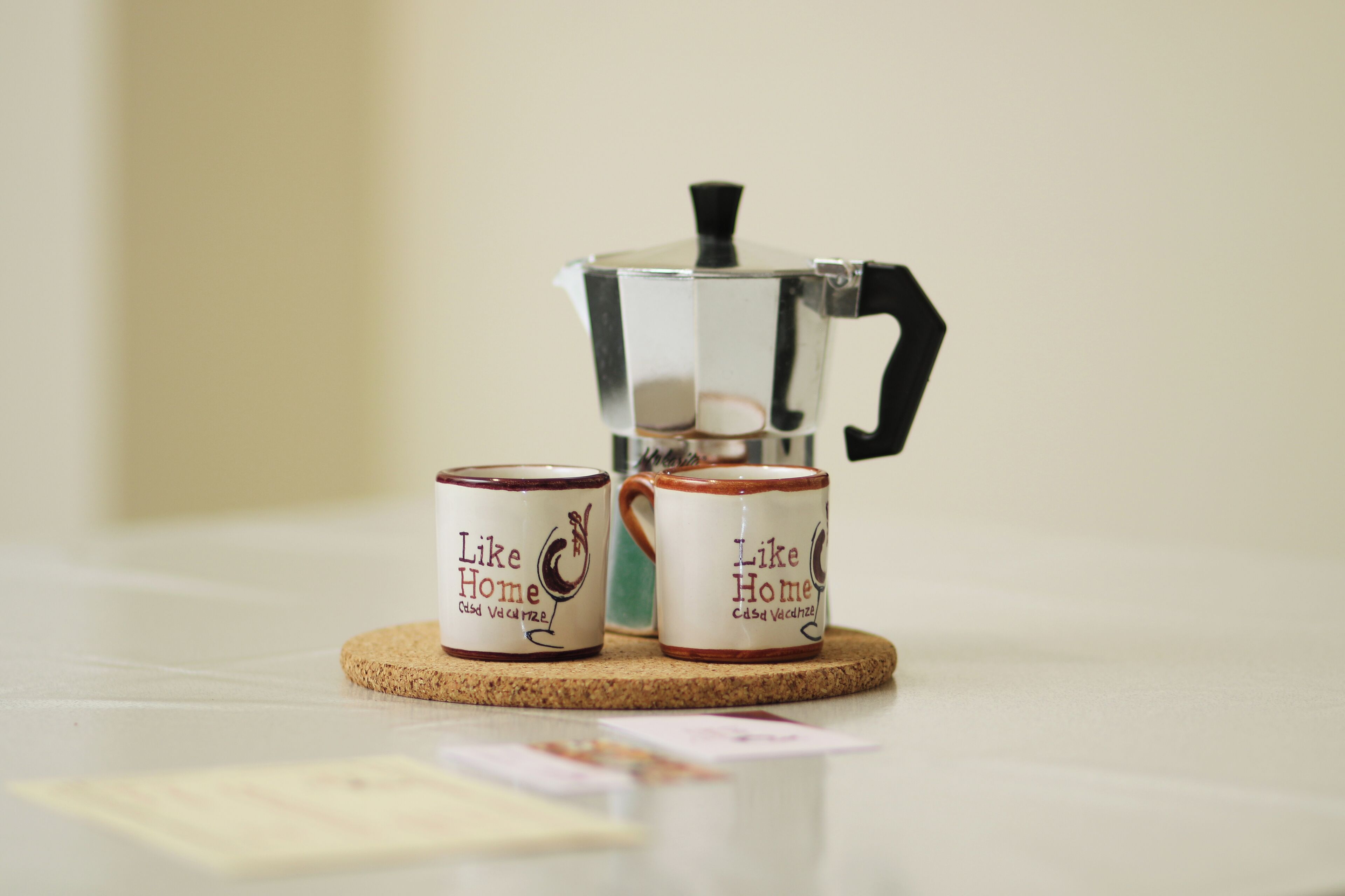 Duas chávenas de café com o texto "Like Home" sobre um apoio de cortiça, uma cafeteira e um cartão desfocado sobre a mesa.