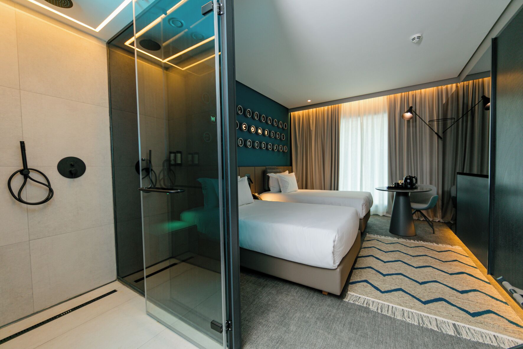 Quarto de hotel moderno com box de vidro, cama com duas almofadas, mesa redonda pequena com cafeteira e painel de controle na parede.