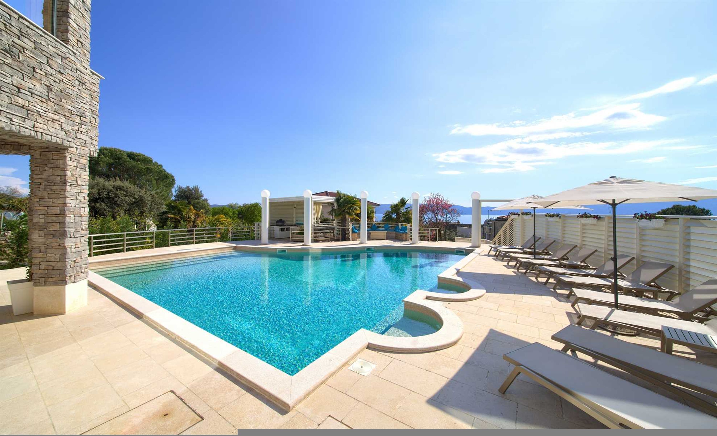 Piscina com espreguiçadeiras e guarda-sóis, cercada por muros de pedra e vegetação.