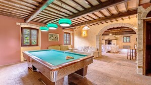 Uma mesa de bilhar em um espaço com vigas de madeira e paredes de pedra, um sofá e uma área de refeições ao fundo.