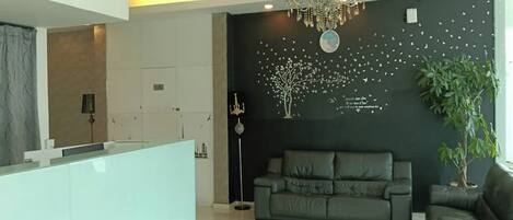 Uma área de recepção moderna com balcão de vidro, parede escura com motivo de árvore e pássaros, lustre e sofá.