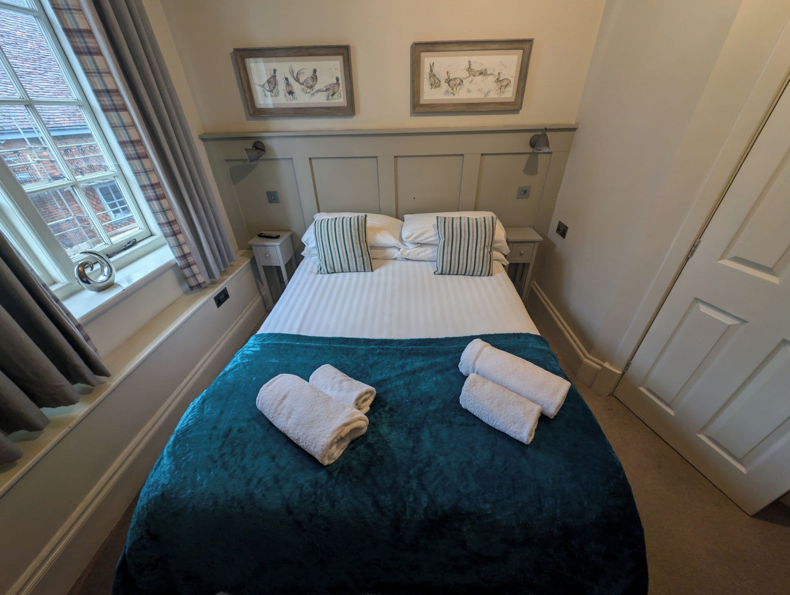 Quarto de hotel com uma cama, duas almofadas, um edredom verde-água, duas toalhas enroladas, uma mesa de cabeceira, um abajur e uma janela com cortinas. Há também duas molduras com quadros na parede.
