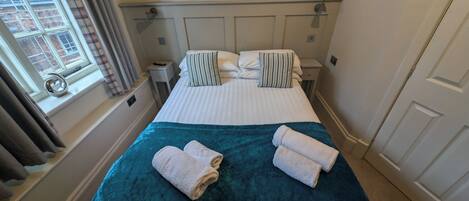 Quarto de hotel com uma cama, duas almofadas, um edredom verde-água, duas toalhas enroladas, uma mesa de cabeceira, um abajur e uma janela com cortinas. Há também duas molduras com quadros na parede.