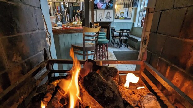 Uma lareira com lenha acesa, um espaço de bar com bancos e balcão, e uma área de estar com mesas e cadeiras.