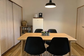 Uma área de refeições com mesa de madeira, cadeiras pretas, luminária pendente, geladeira branca e prateleira com utensílios de cozinha.