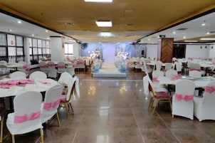 Salão de festas com mesas redondas, cadeiras brancas e uma passagem central que leva a um palco.