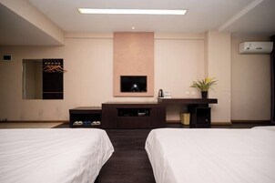 Quarto de hotel com duas camas, uma televisão fixada na parede, uma secretária pequena com uma planta e um porta-sapatos.