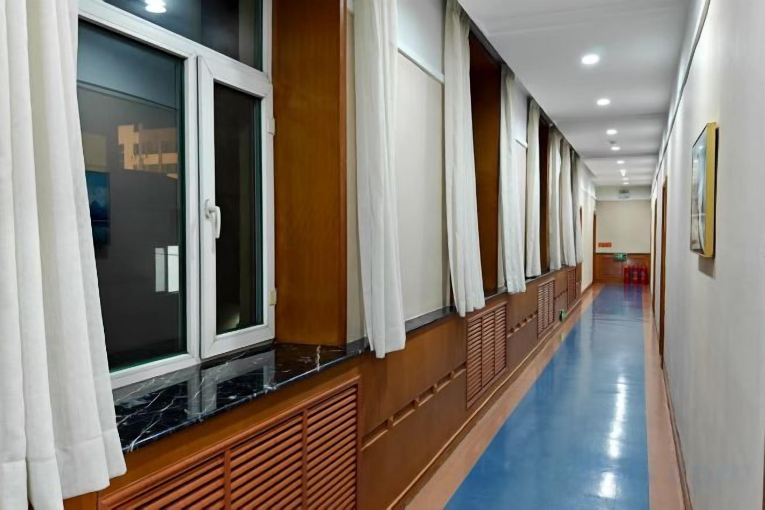 Corredor de hotel com chão azul, painéis de madeira e janela com cortinas.
