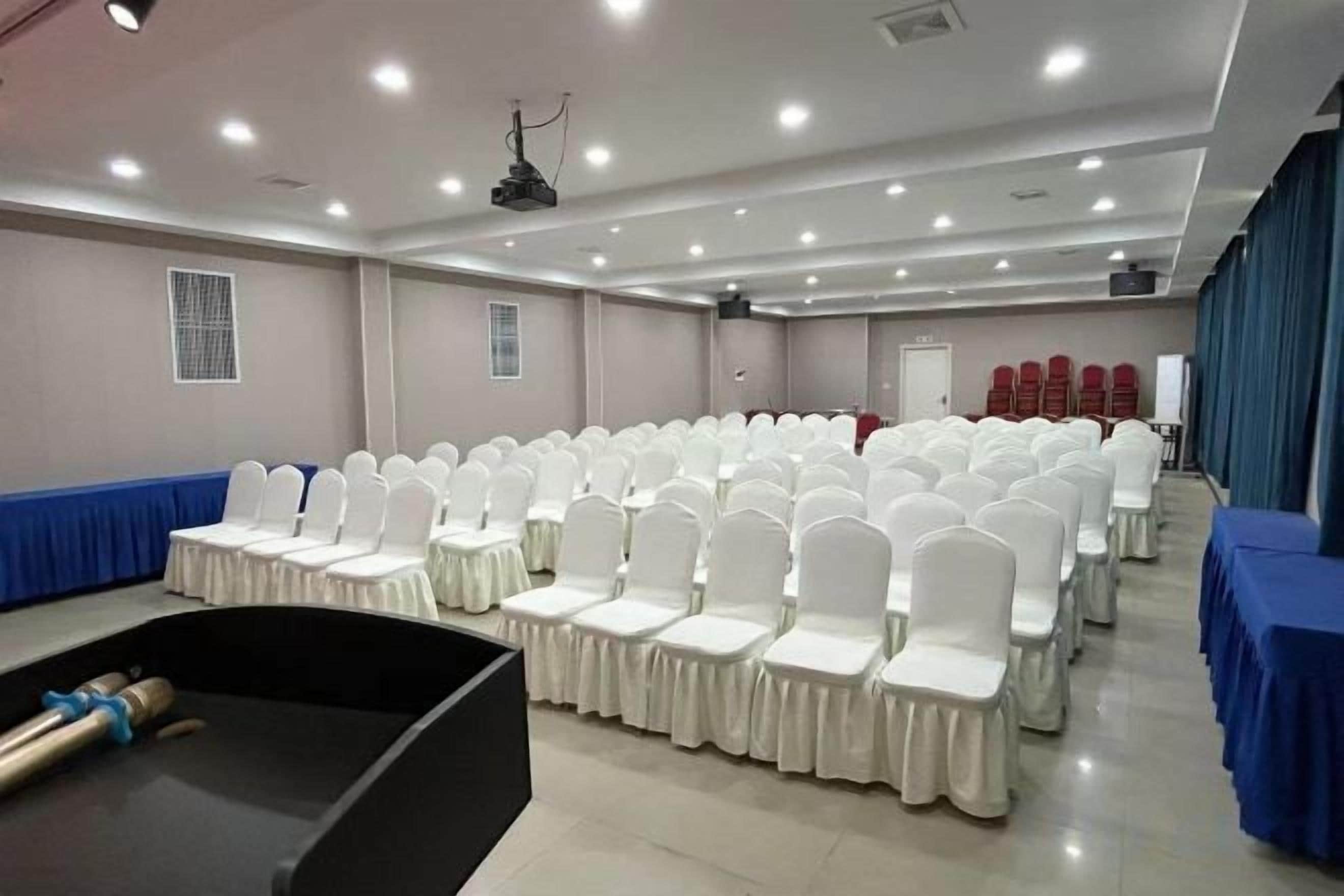 Sala de conferência com fileiras de cadeiras brancas, um palco e uma mesa azul à direita.