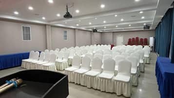 Sala de conferência com fileiras de cadeiras brancas, um palco e uma mesa azul à direita.