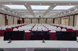 Uma sala de conferências com fileiras de cadeiras brancas, uma mesa comprida com toalha vermelha e um teto com claraboias.