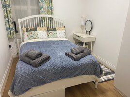Cama bem arrumada com colcha azul e duas toalhas dobradas, mesa de cabeceira branca com espelho e uma pequena escrivaninha com abajur.