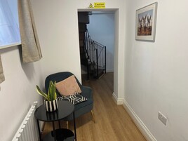 Um corredor com escadas, uma cadeira preta com almofada, uma pequena mesa com uma planta e um quadro na parede.